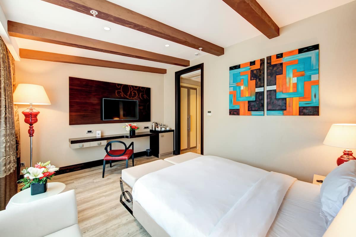 Quarto de hotel com cama grande, escrivaninha com cadeira, televisão e obras de arte nas paredes.
