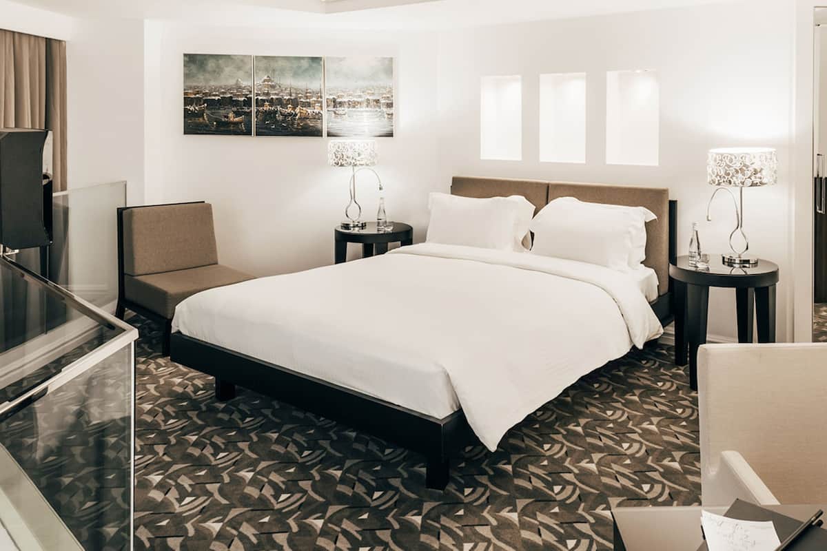 Quarto de hotel com cama grande, duas mesas de cabeceira, uma cadeira, escrivaninha, televisão e pintura com vista para a cidade.