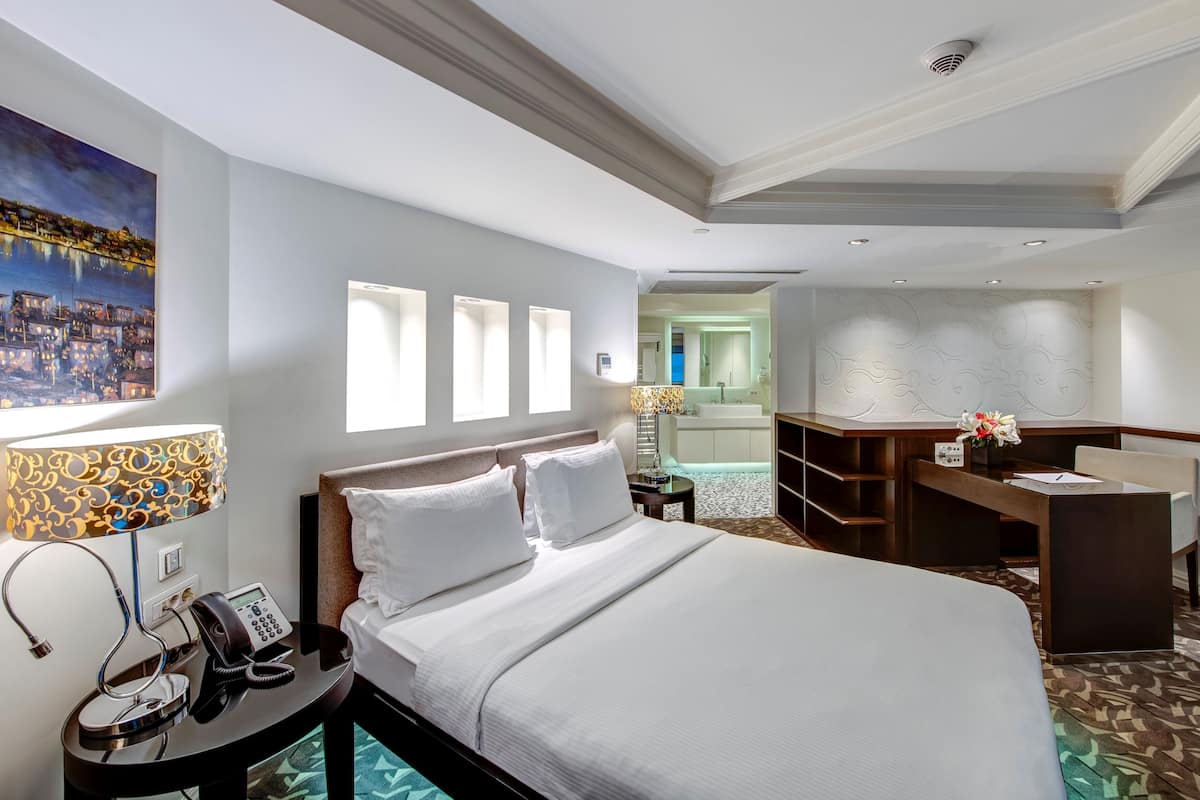 Quarto de hotel com cama grande, mesa de cabeceira com abajur, escrivaninha com cadeira, banheiro com espelho e um quadro na parede.