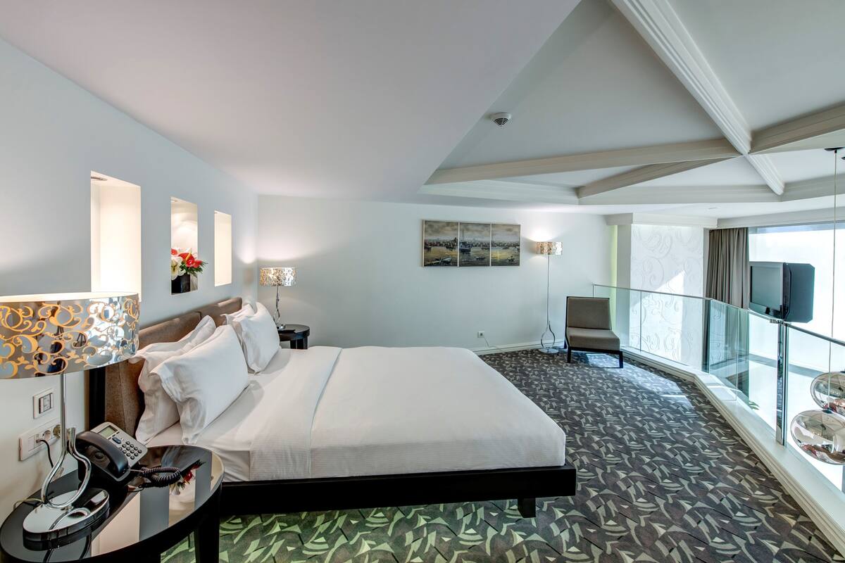 Quarto de hotel com uma cama grande, luminárias laterais, telefone, um quadro na parede, uma cadeira e uma varanda com guarda-corpo de vidro.