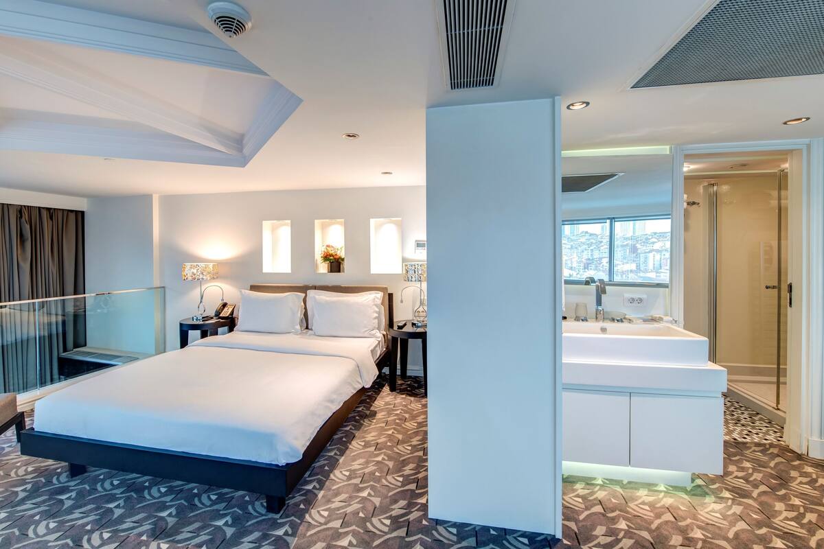 Quarto de hotel moderno com uma cama grande, uma escrivaninha, uma cadeira, uma mesinha, um abajur, um espelho, uma janela e um banheiro com banheira e pia.
