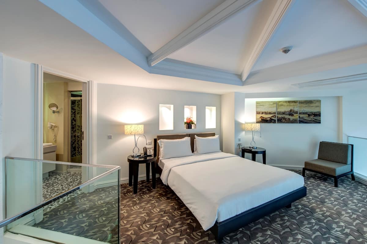Quarto de hotel com uma cama grande, uma cadeira, uma mesa pequena e uma divisória de vidro.