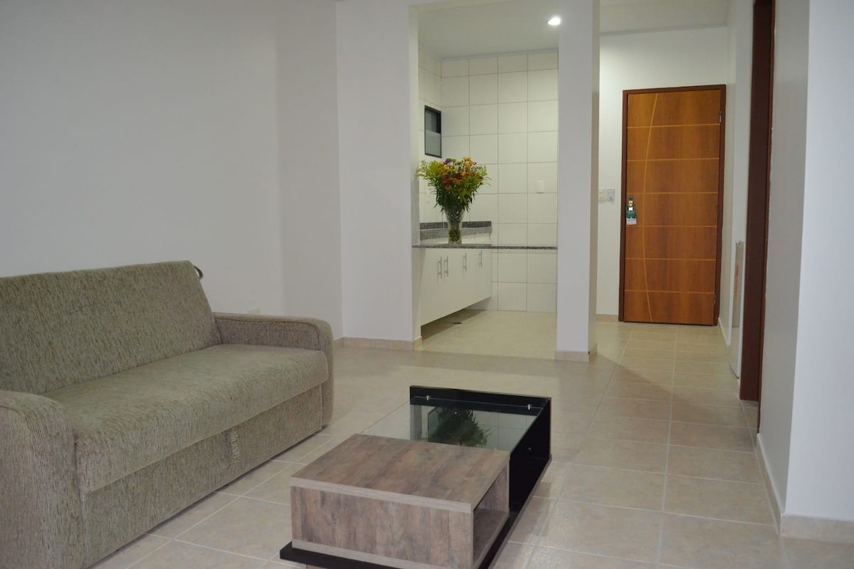 Sala de estar com sofá, mesinha de centro e cozinha visível por uma porta aberta.