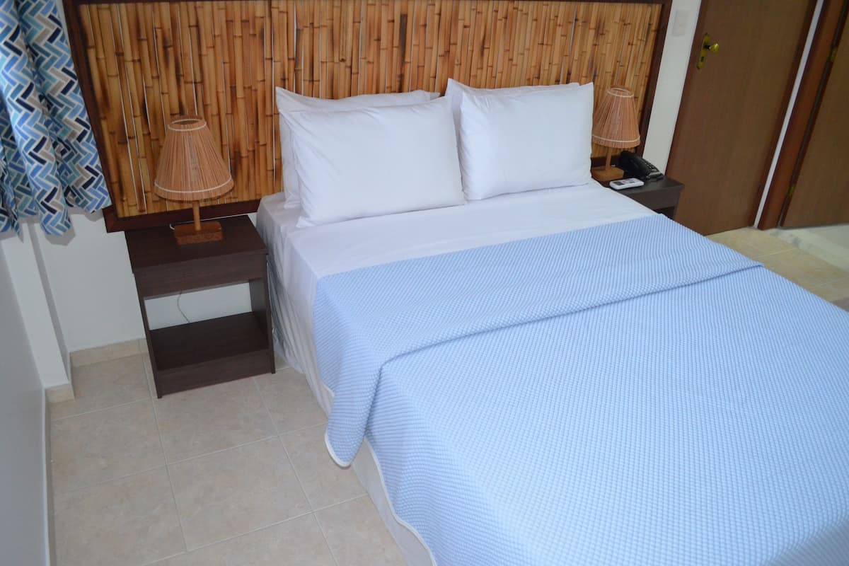 Cama bem arrumada com lençóis brancos e manta listrada em azul, cabeceira de madeira, luminária de cabeceira e mesa de cabeceira com telefone e luminária.