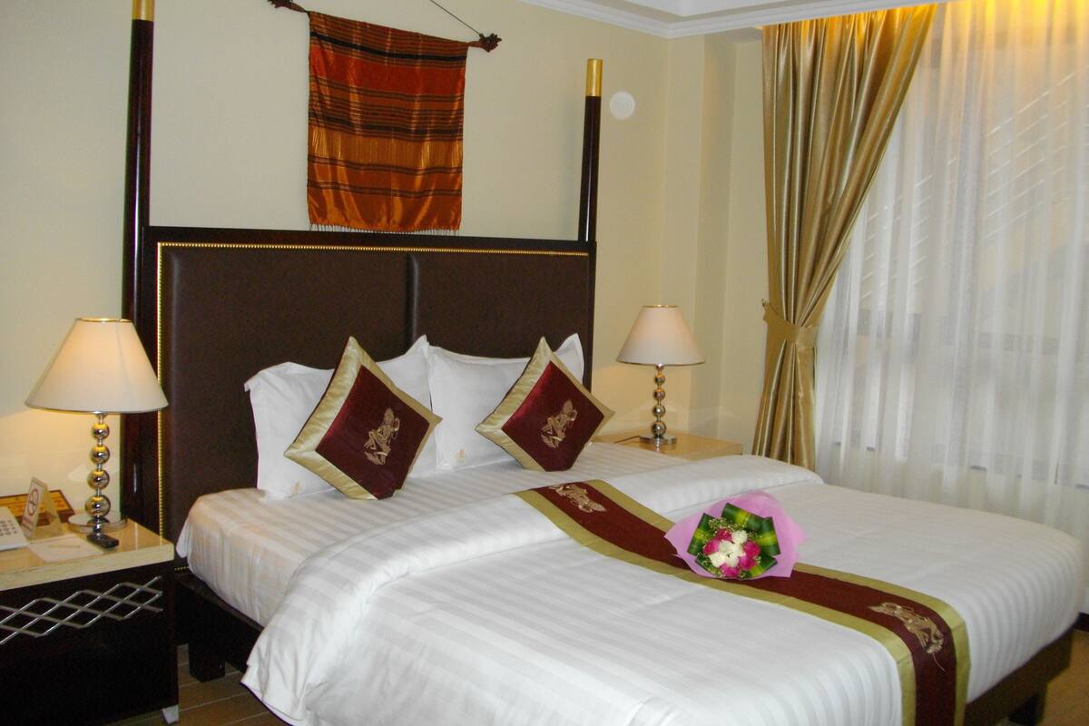 Quarto de hotel com uma cama grande, duas mesas de cabeceira, luminárias, um painel decorativo na cabeceira, um objeto pendurado na parede e uma janela com cortinas.