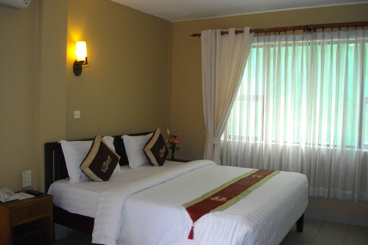 Quarto de hotel com cama, mesa de cabeceira, luminária e janela com cortinas.