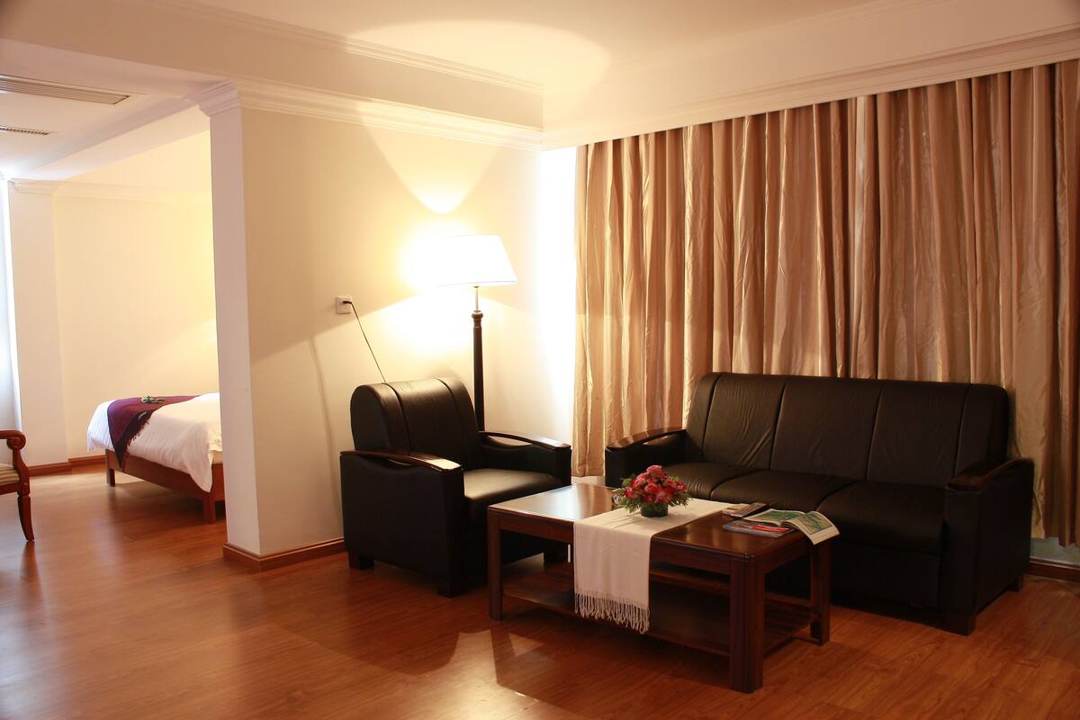 Quarto de hotel com cama, duas poltronas de couro preto, mesa de centro de madeira, abajur e uma janela grande com cortinas.