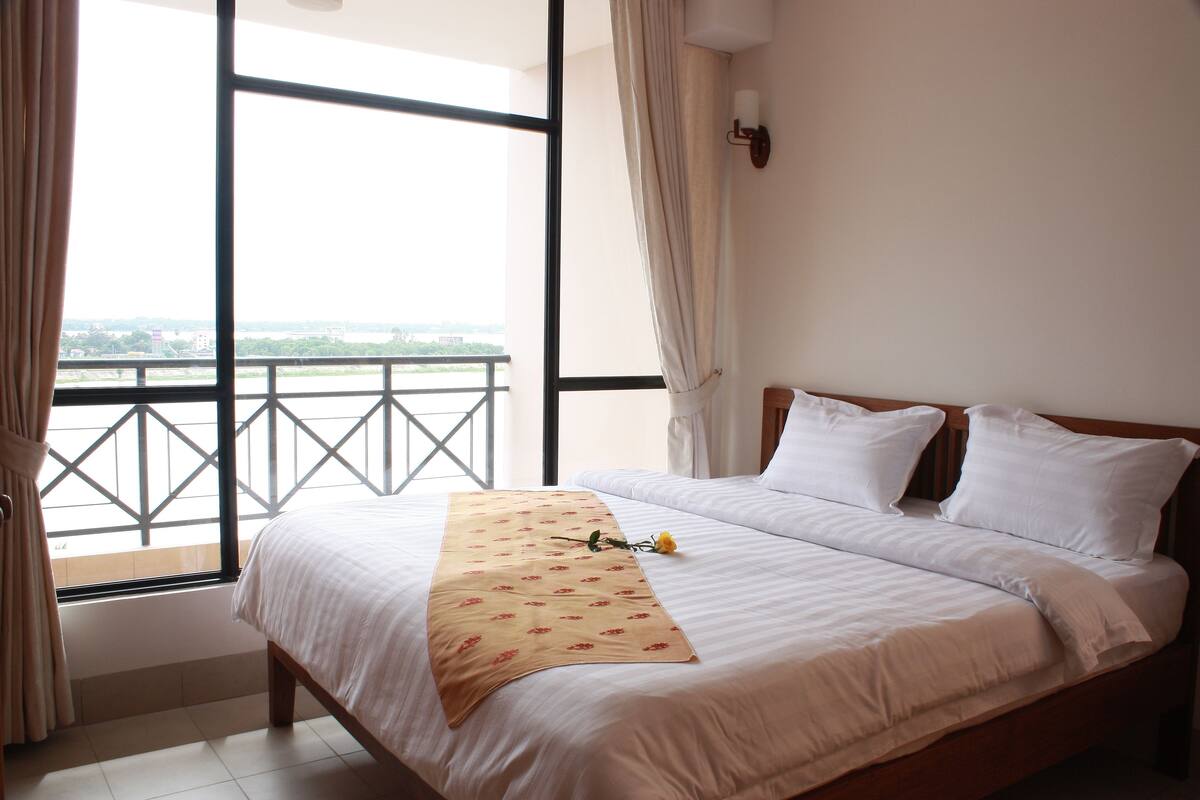 Quarto de hotel com uma janela grande, cama com roupa de cama branca e manta de banquinho com padrão floral.