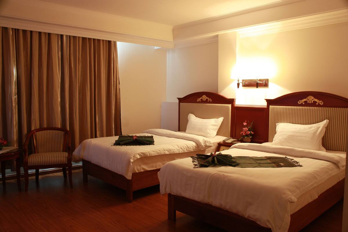 Quarto de hotel com duas camas, uma cadeira, uma mesa de cabeceira e uma luminária.