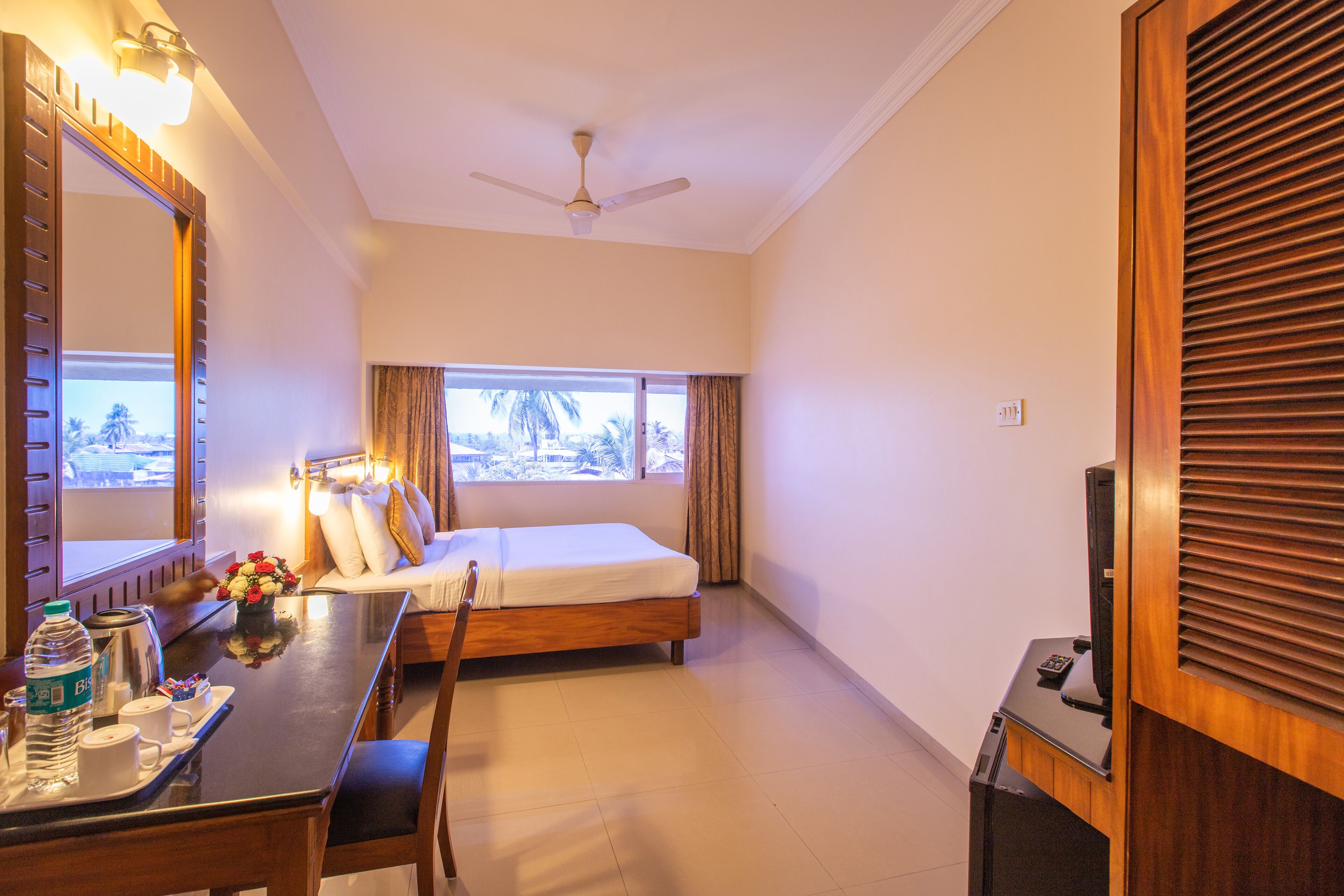 Quarto de hotel com cama, escrivaninha com espelho, televisão e janela com cortinas.
