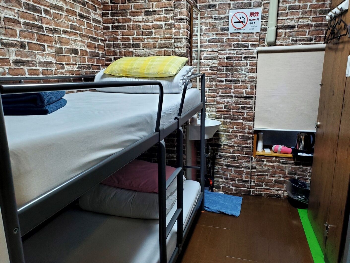 Quarto com beliche, parede de tijolos, placa de proibido fumar, pia e pequena prateleira com pertences pessoais.