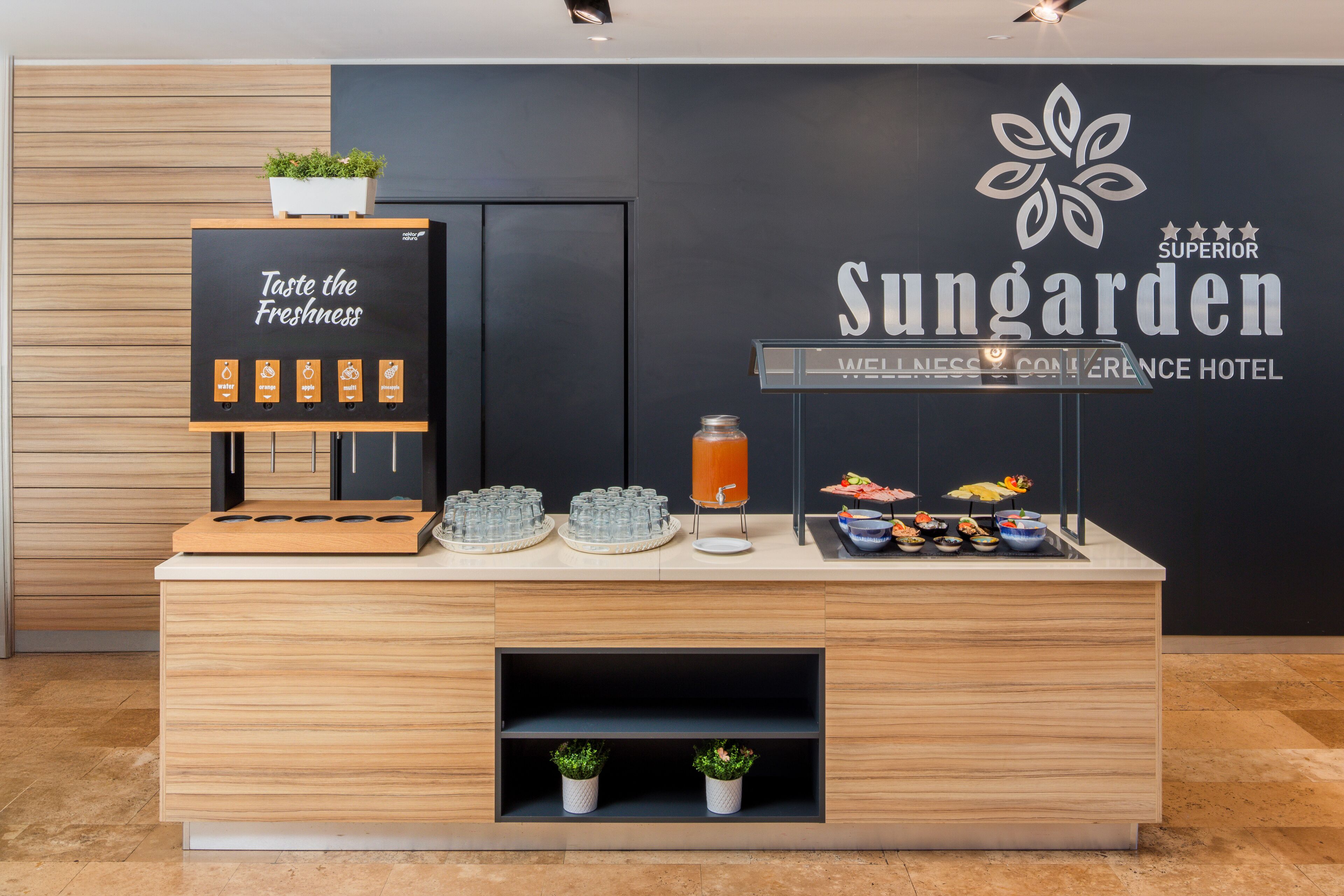 Uma área de recepção com balcão de madeira, um letreiro com a inscrição "Sungarden Wellness Conference Hotel" e uma variedade de frutas e bebidas frescas.