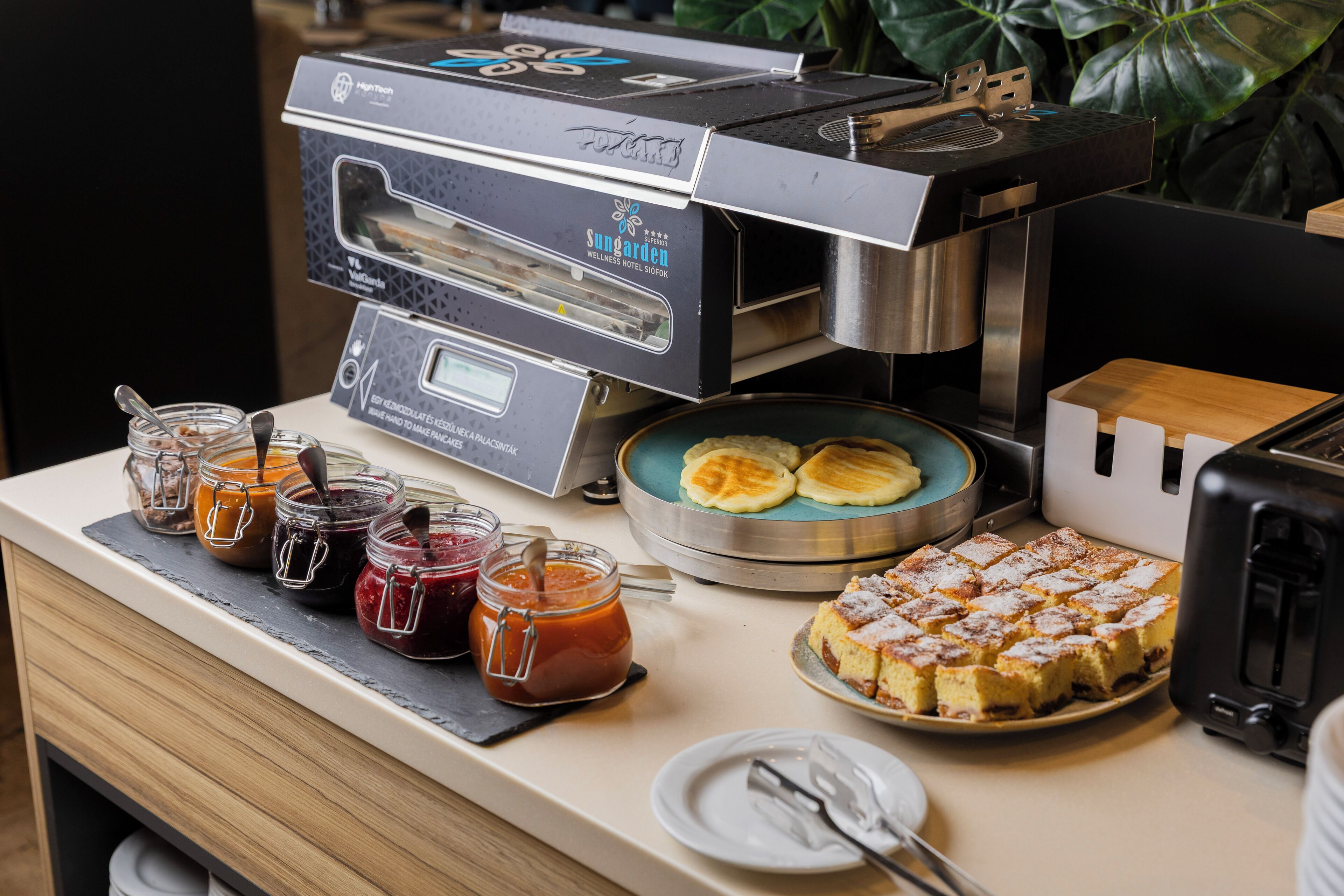 Disposição de buffet de café da manhã com máquina de café, diversas geleias, um prato de panquecas e uma fatia de bolo.