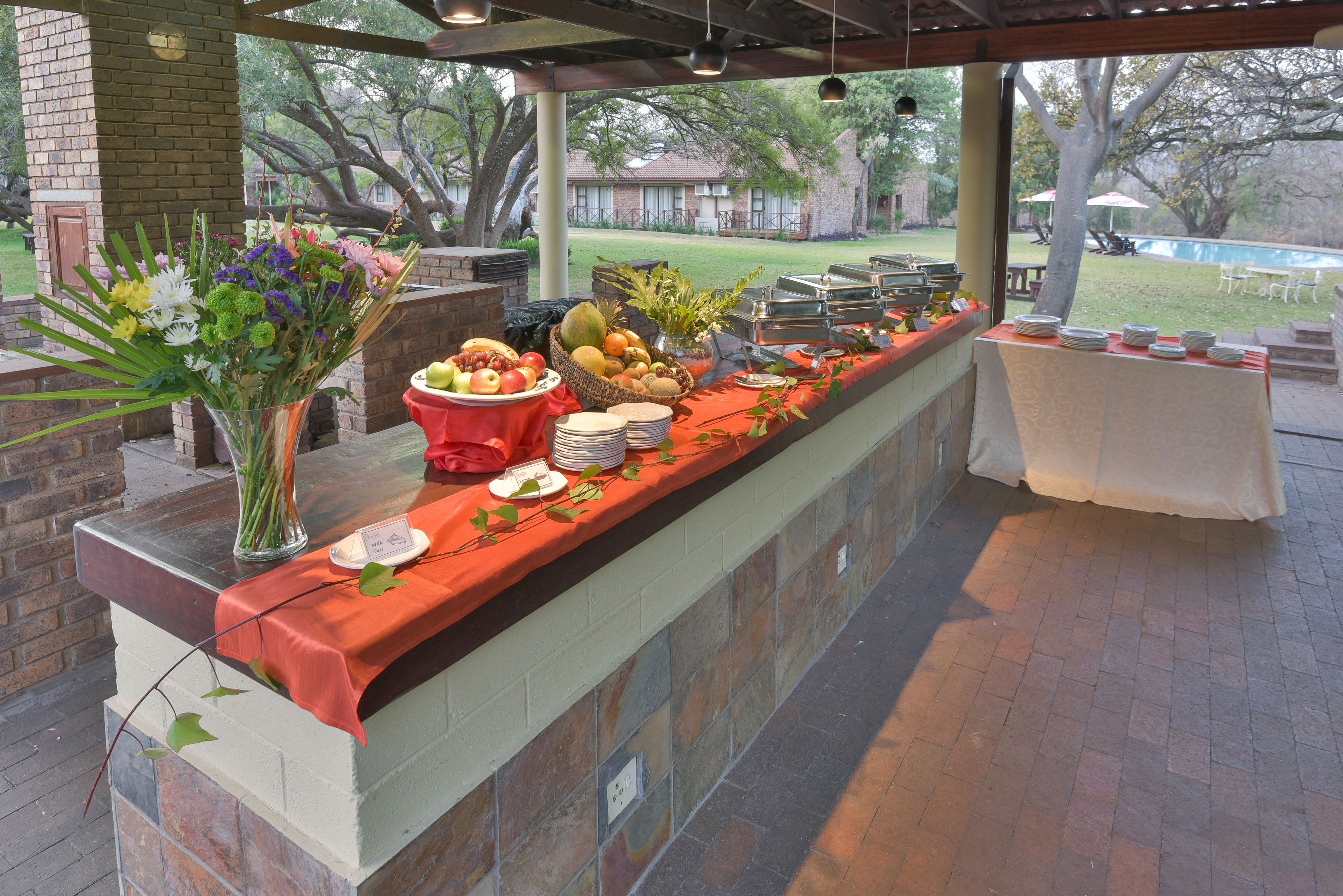 Buffet ao ar livre com diversas frutas e legumes, um vaso de flores e uma mesa com pratos e guardanapos.