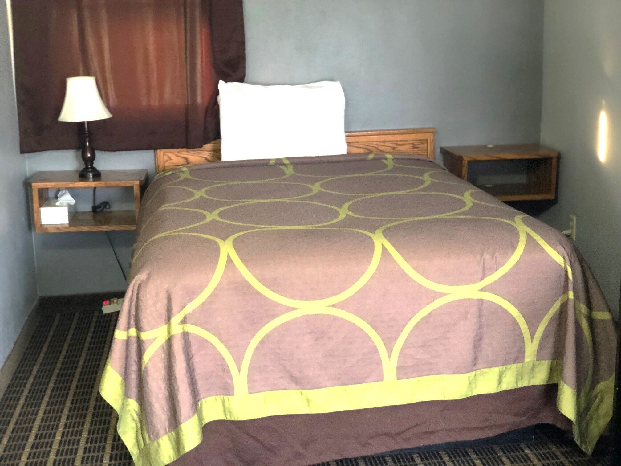 Quarto de hotel com cama, mesa de cabeceira com abajur, cabeceira de madeira e pequena prateleira.