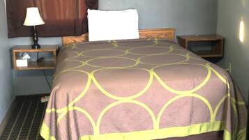Quarto de hotel com cama, mesa de cabeceira com abajur, cabeceira de madeira e pequena prateleira.