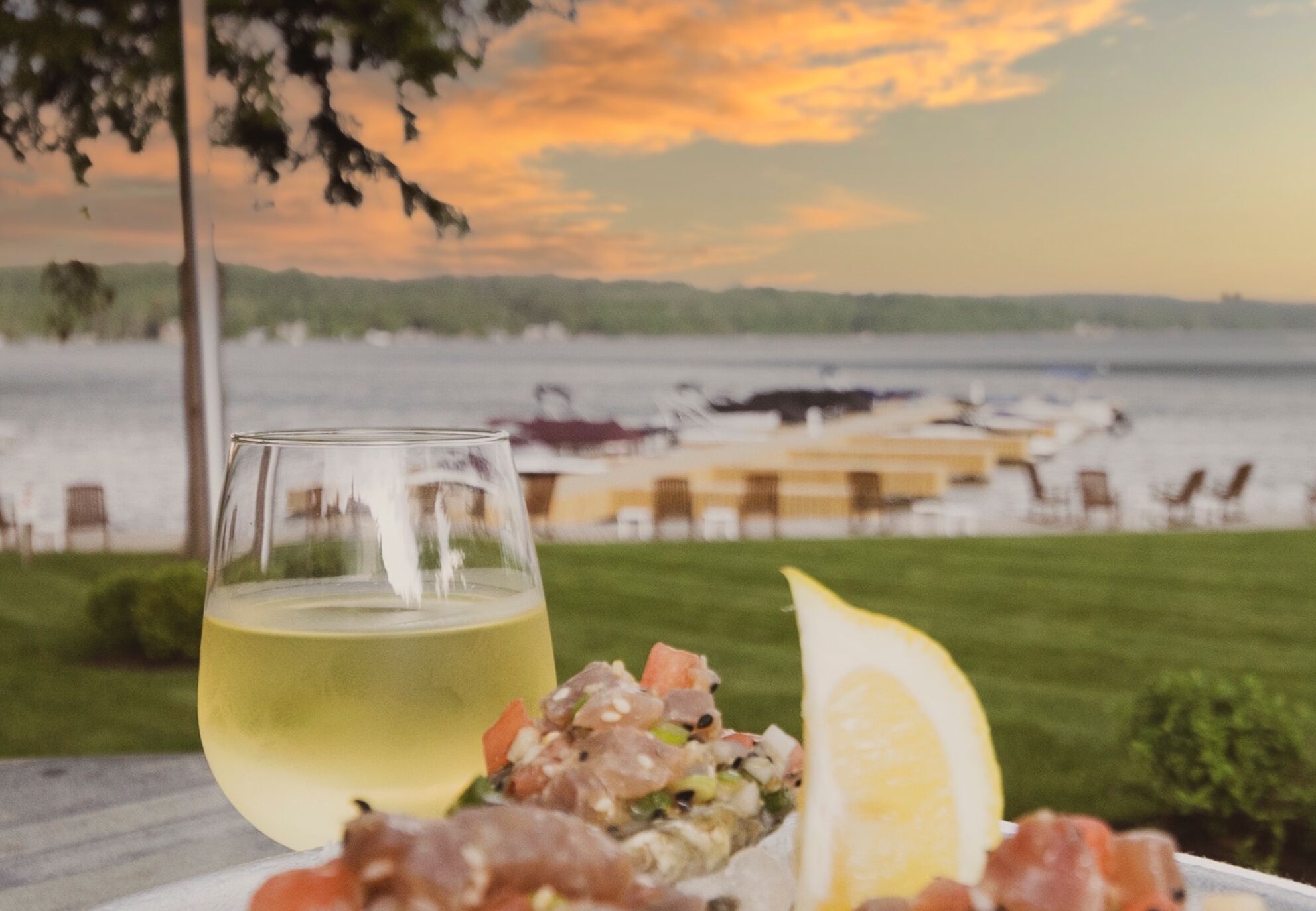 Um copo de vinho branco, metade de um limão e um prato de frutos do mar sobre uma mesa, com vista para um lago e barcos ao fundo.