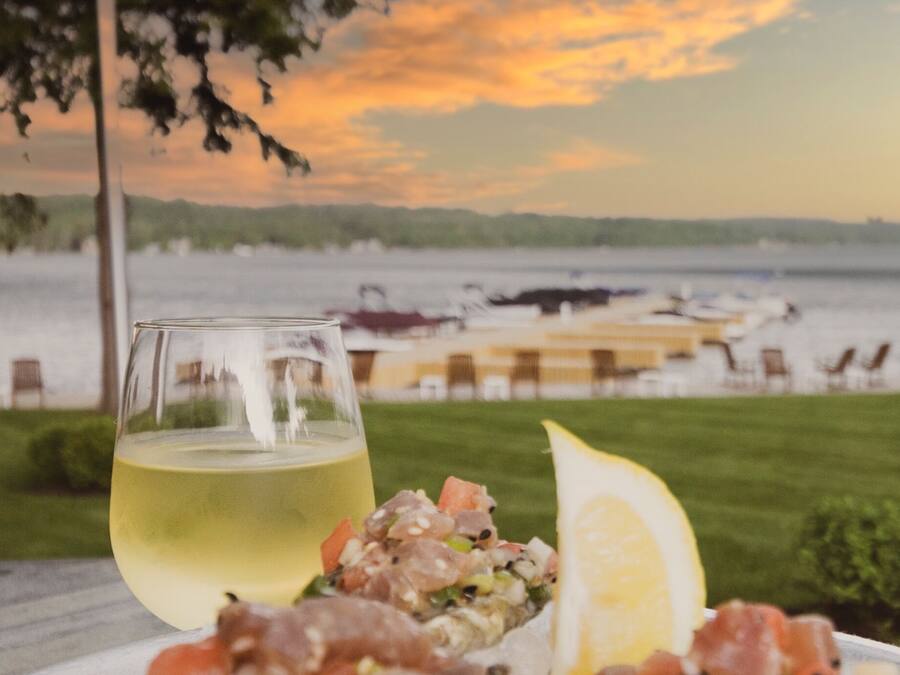 Um copo de vinho branco, metade de um limão e um prato de frutos do mar sobre uma mesa, com vista para um lago e barcos ao fundo.