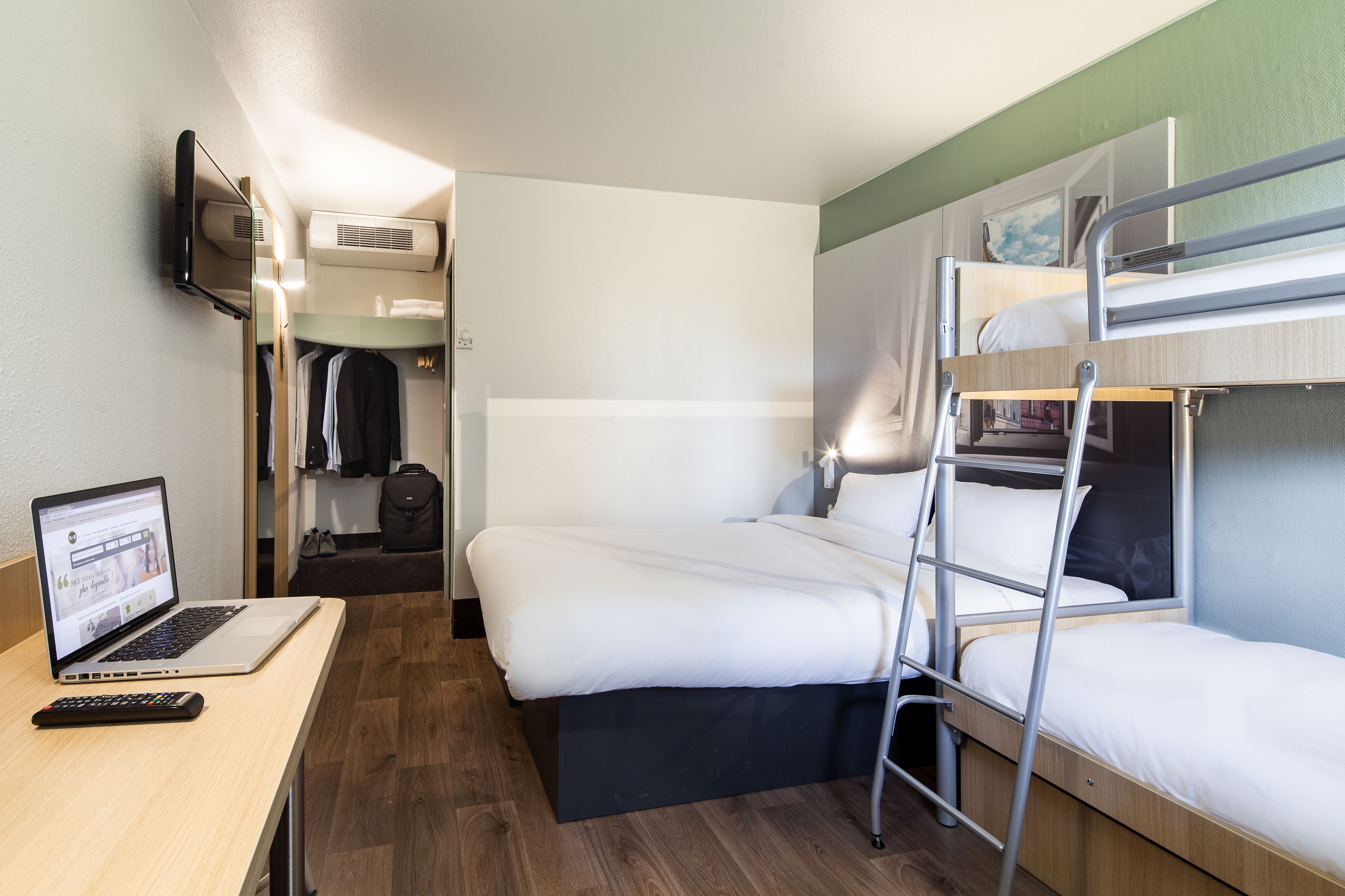 Quarto de hotel pequeno com uma cama beliche, uma escrivaninha com um laptop e um espelho.
