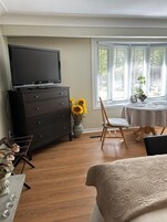 Quarto com televisão em uma cômoda, mesa com cadeira e janela com persianas.
