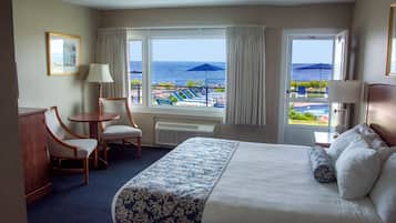 Quarto de hotel com uma cama, duas cadeiras, uma mesinha e uma janela com vista para o mar.