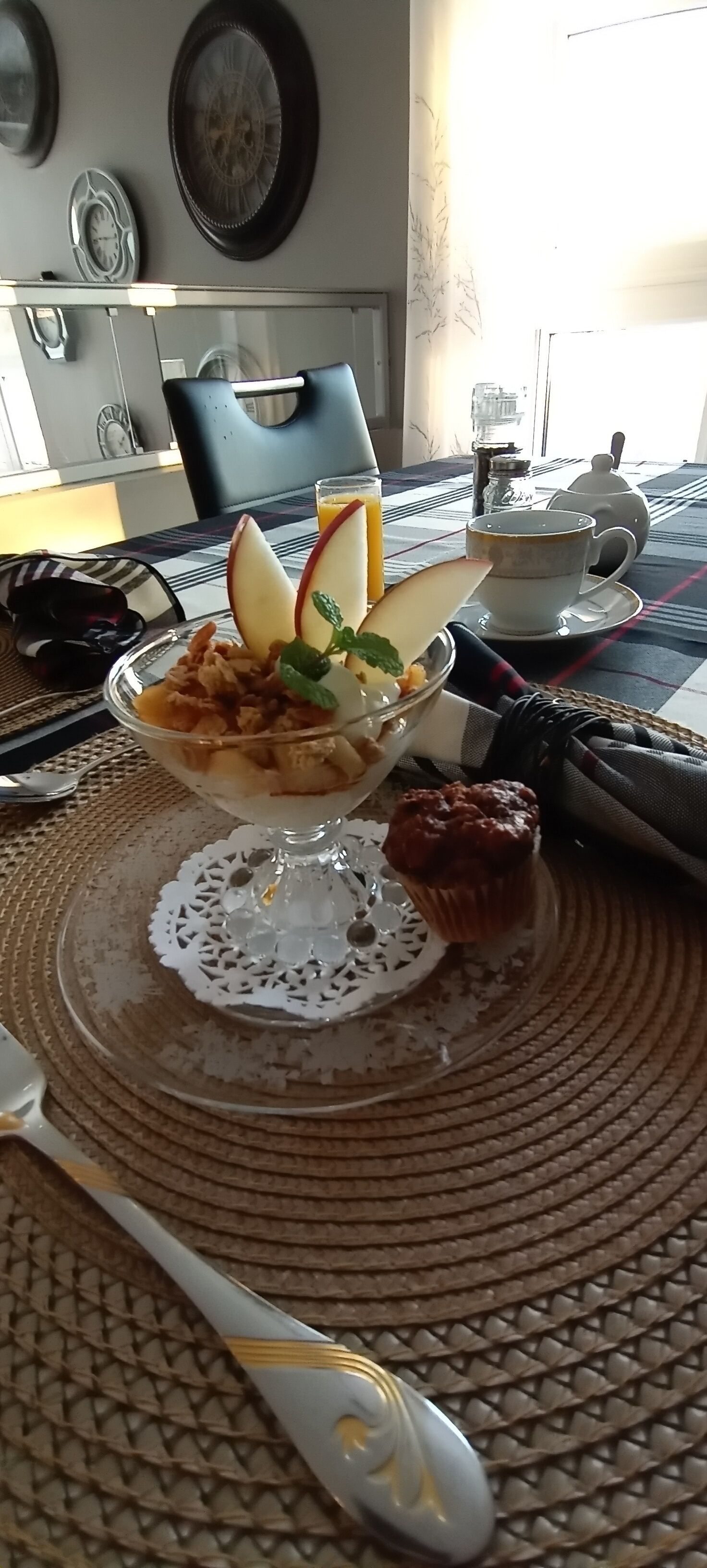 Disposição para o café da manhã com uma tigela de cereal coberto com maçãs e hortelã, um muffin, uma xícara e uma chaleira sobre uma toalha xadrez.