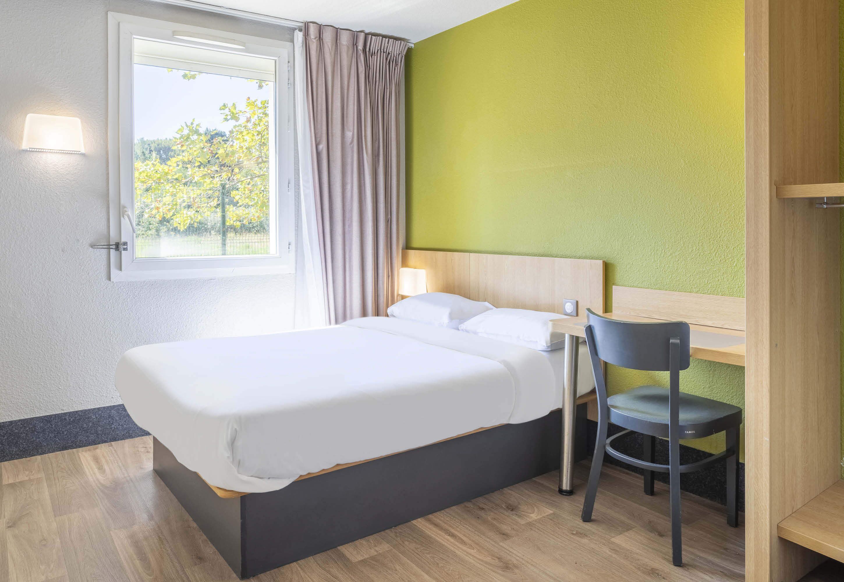 Quarto de hotel com cama, escrivaninha, cadeira, janela com cortinas e luminária.