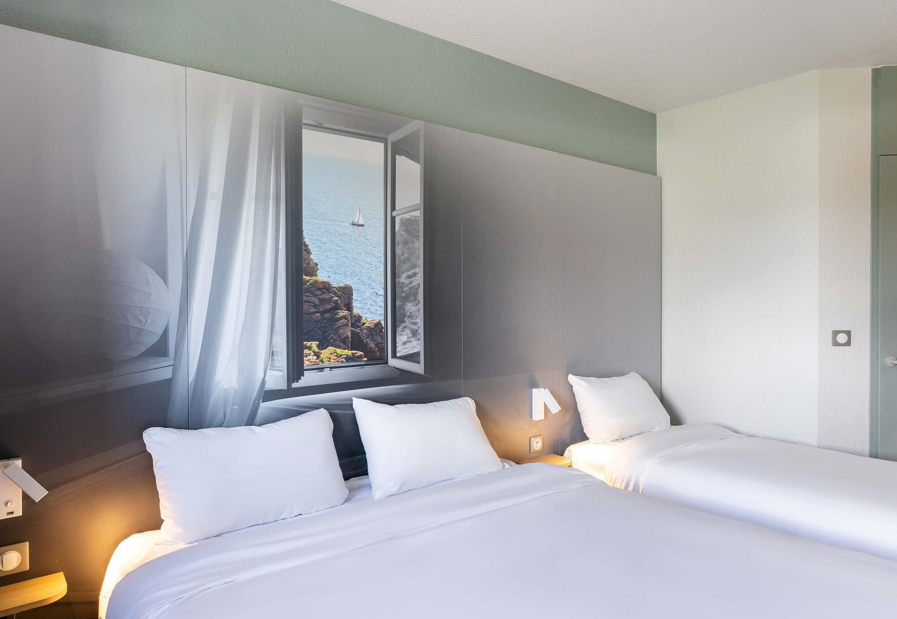 Quarto de hotel com cama, mesa de cabeceira, janela com vista para o mar e luminária na parede.