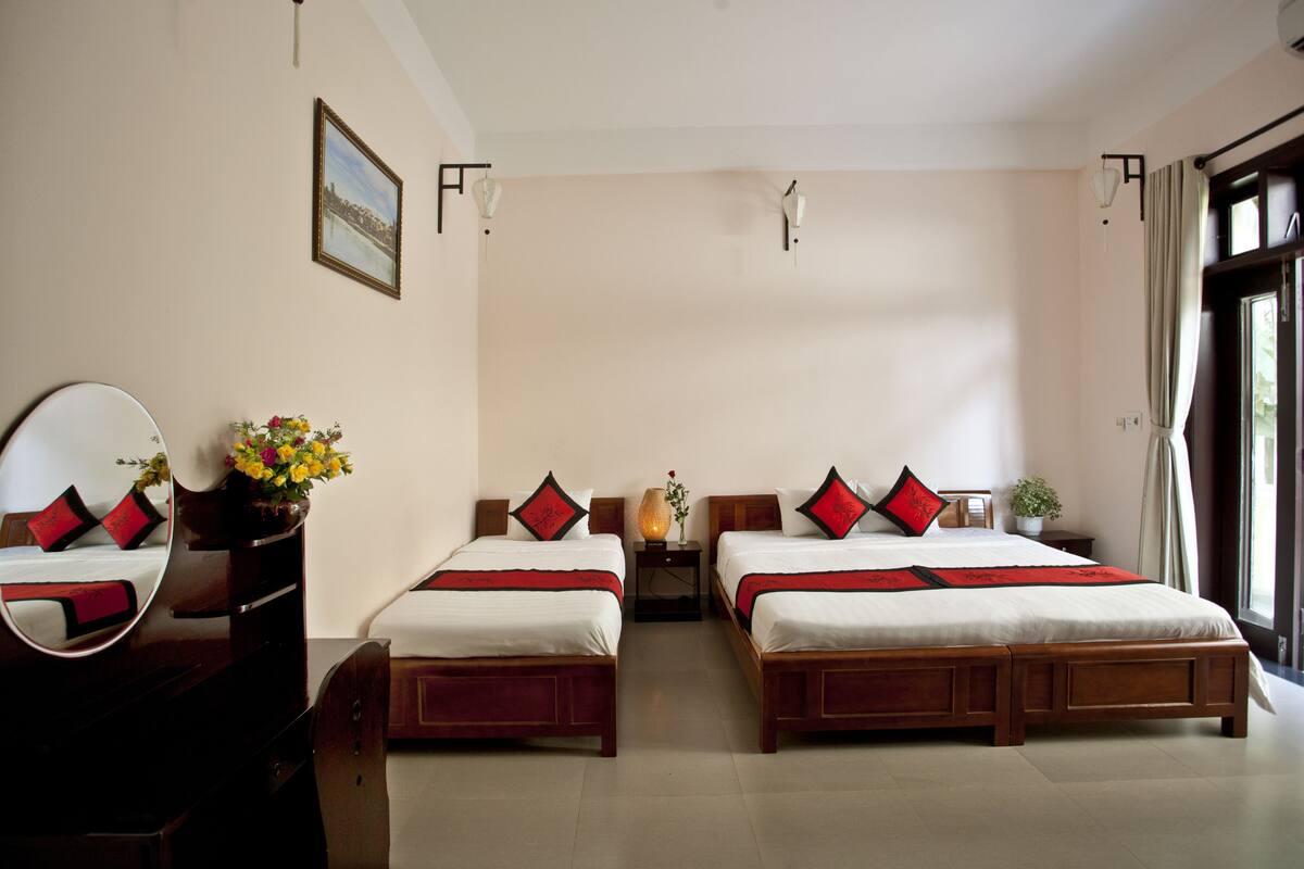 Quarto de hotel com duas camas, uma cômoda, um espelho, um vaso com flores e uma janela com cortinas.