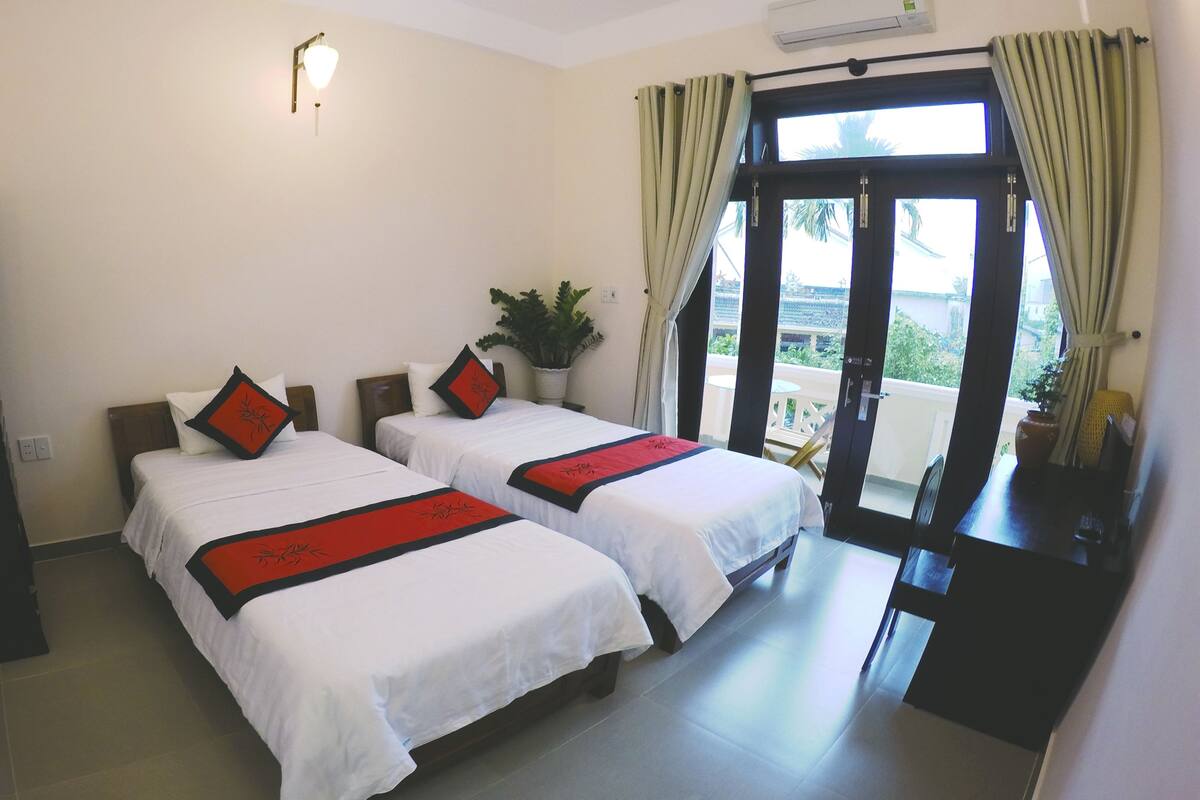 Quarto de hotel com duas camas, um vaso de planta e vista para o exterior através de uma porta.