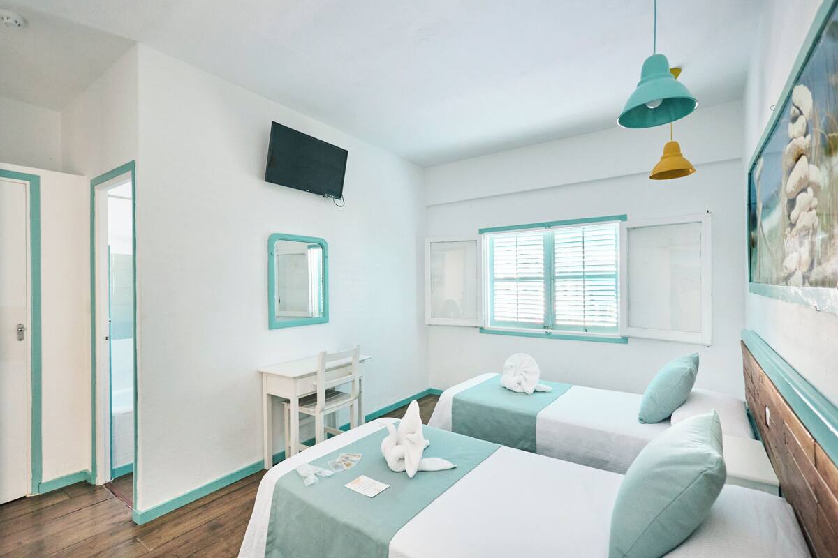 Quarto com duas camas, uma escrivaninha, uma cadeira, uma televisão e uma janela com persianas.