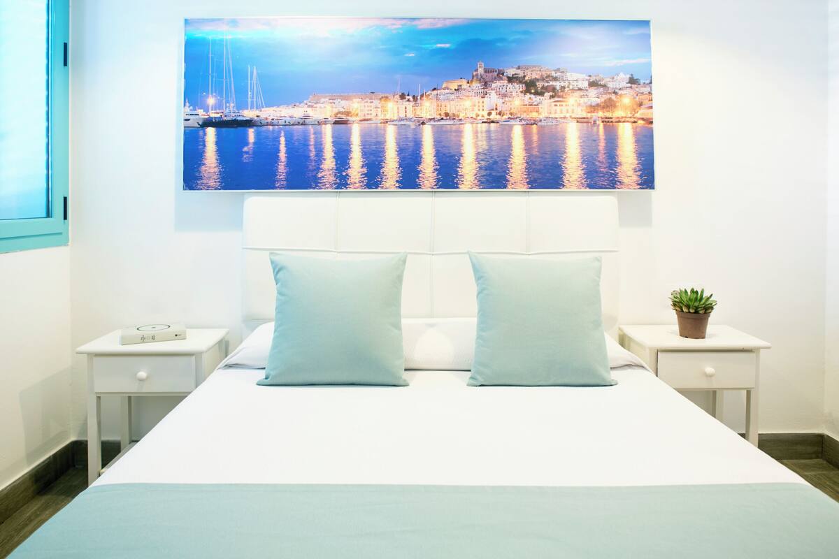 Quarto de hotel com cama, duas almofadas, mesa de cabeceira com uma planta e um quadro na parede retratando uma paisagem costeira.