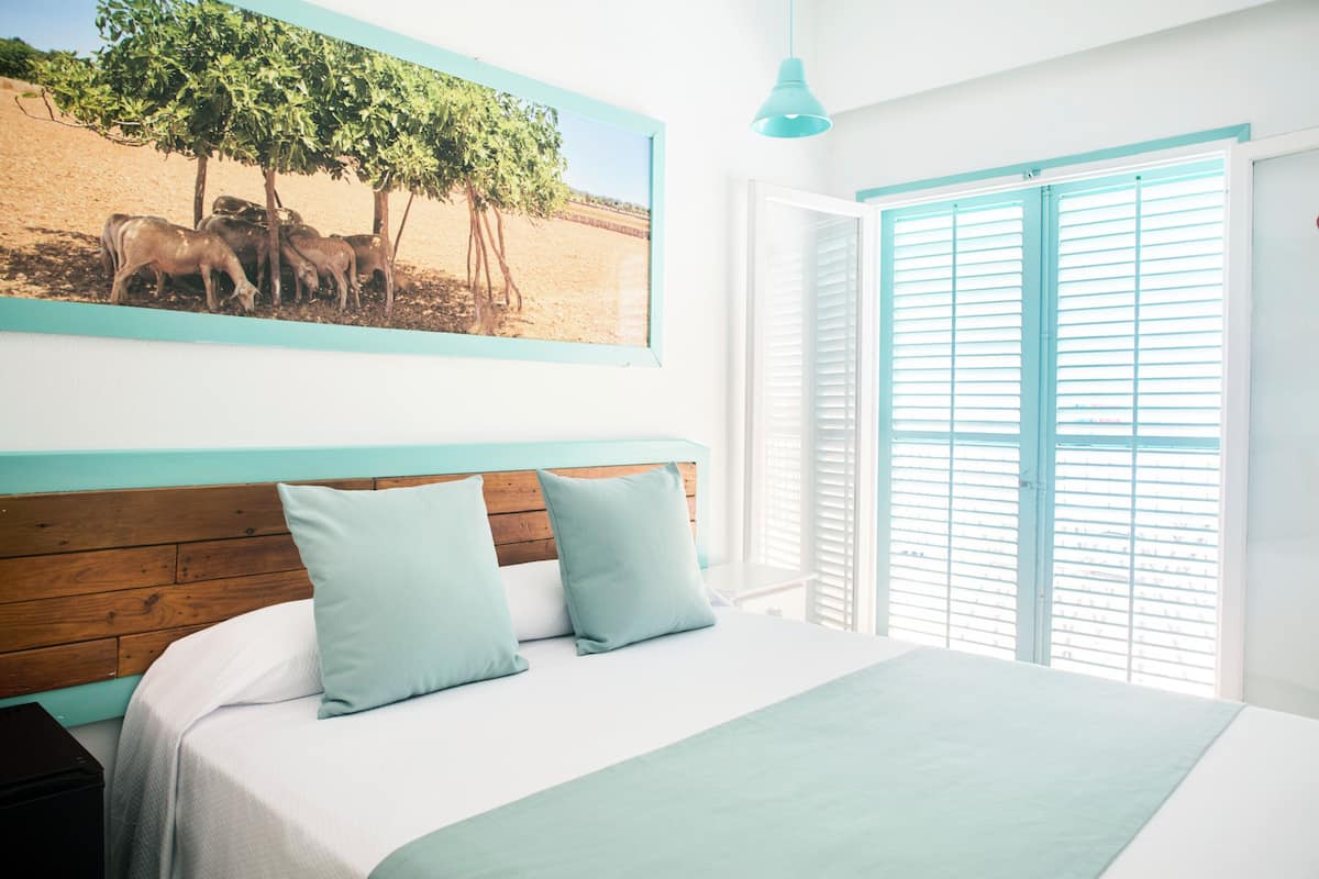 Um quarto com cama, mesa de cabeceira, quadro na parede e janela com persianas.