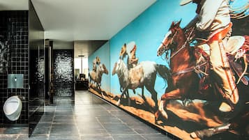 Corredor com um mural de cowboys a cavalo, um mictório e chão de azulejos.