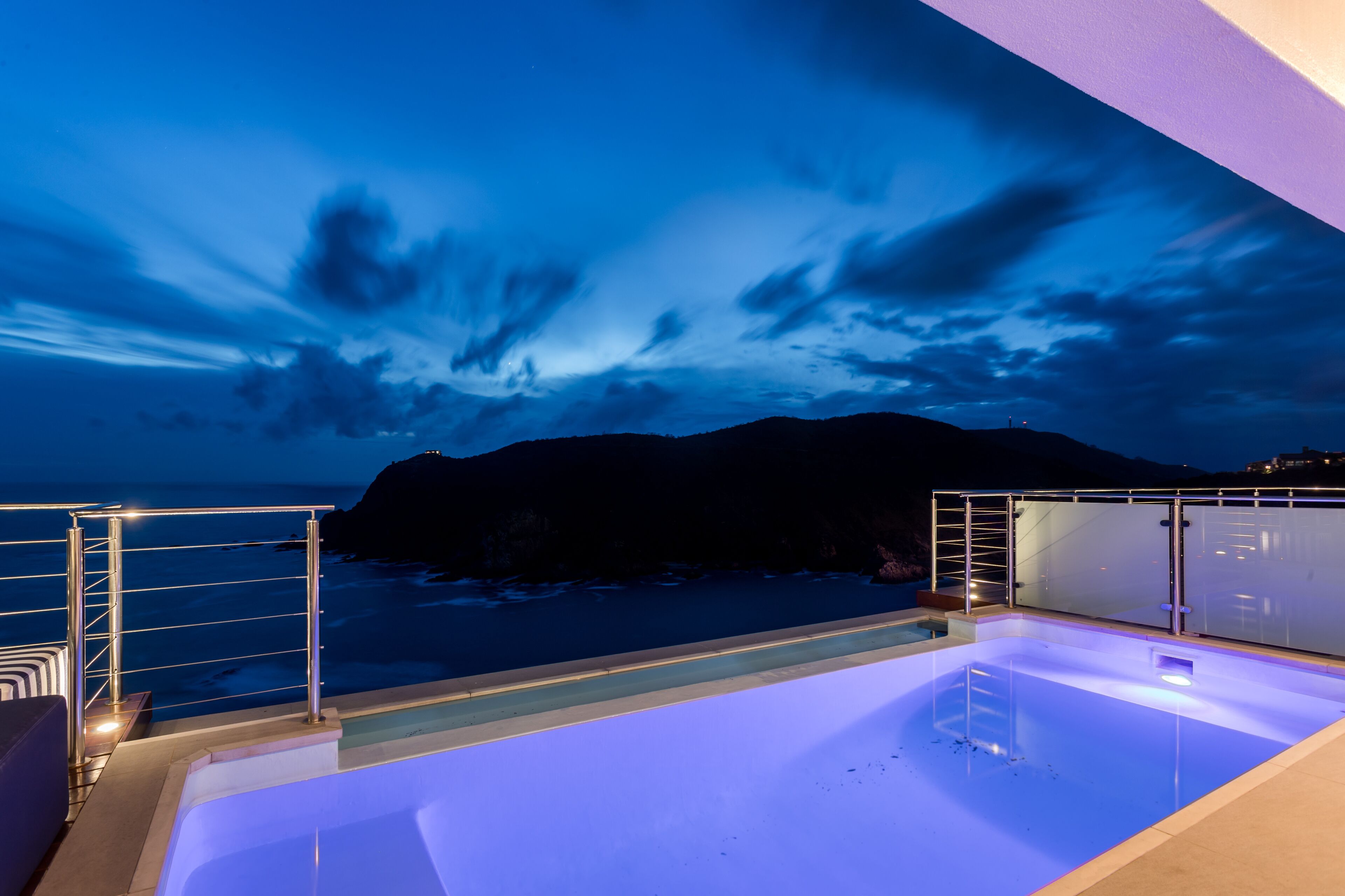 Piscina infinita no terraço, com parapeito de vidro, com vista para a costa montanhosa ao entardecer.