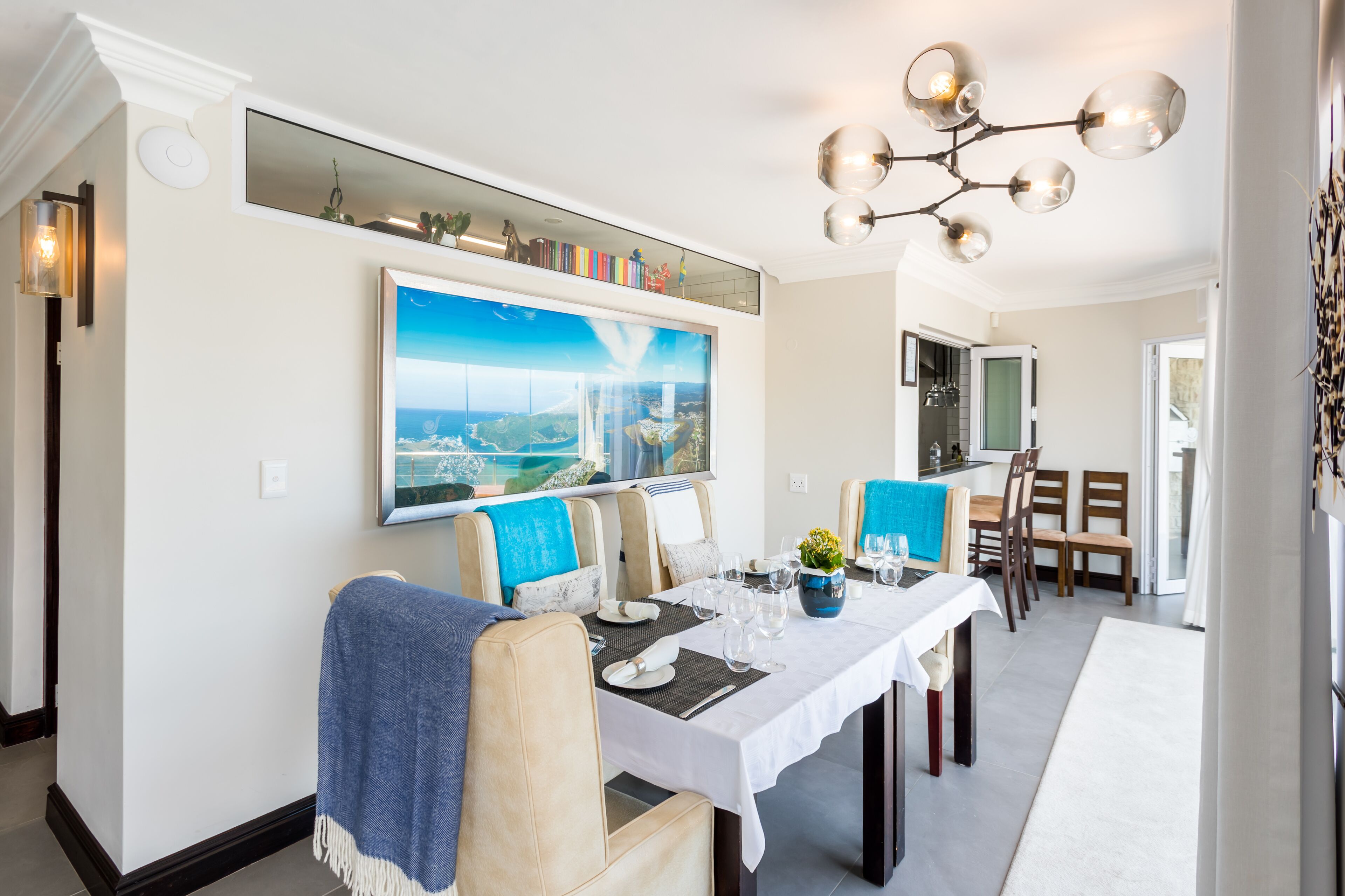 Uma sala de jantar com uma mesa posta para quatro pessoas, um quadro grande acima da mesa e um candelabro.