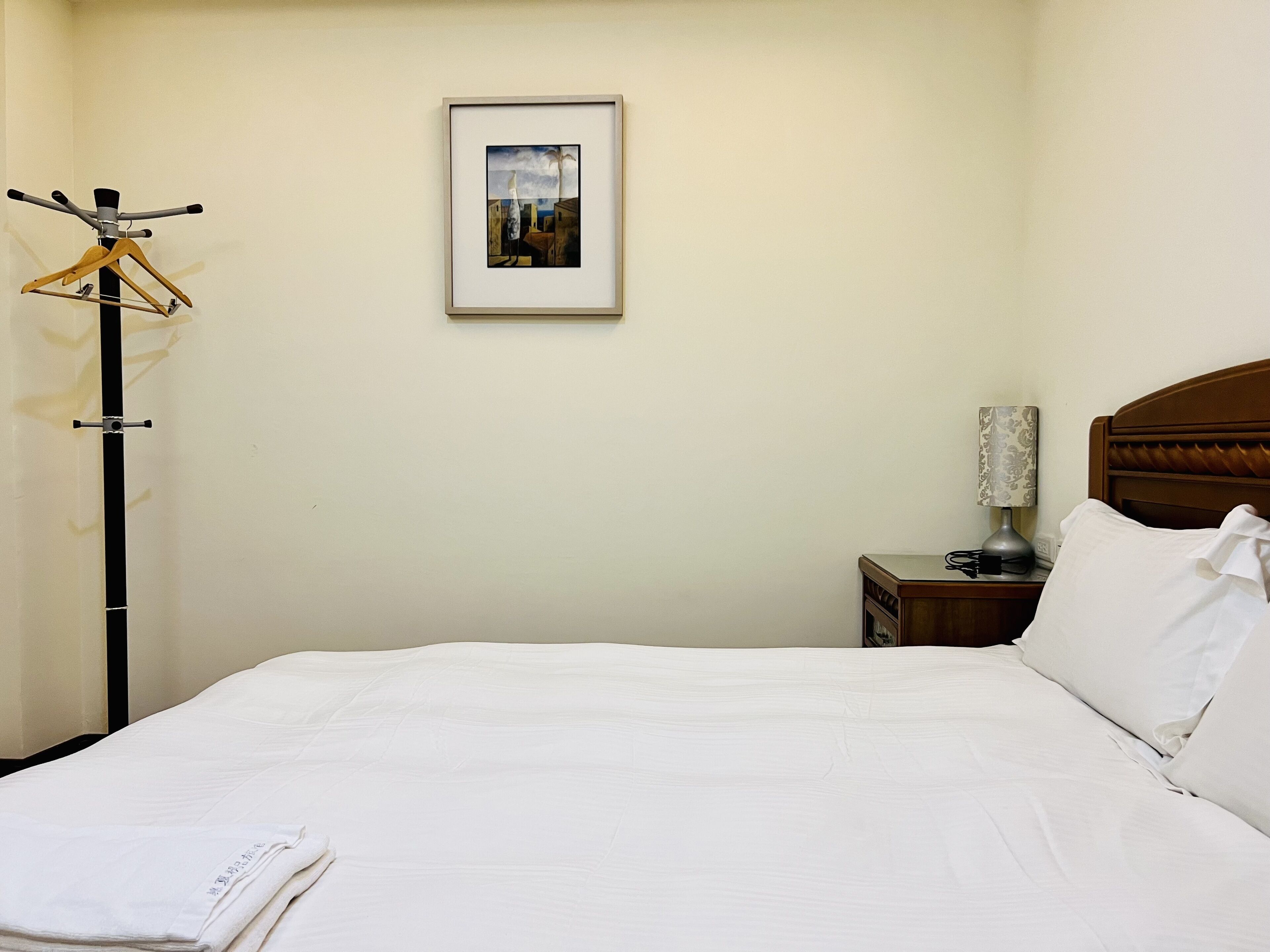 Cama bem arrumada com lençóis brancos, cabeceira de madeira, mesa de cabeceira com abajur e quadro na parede.