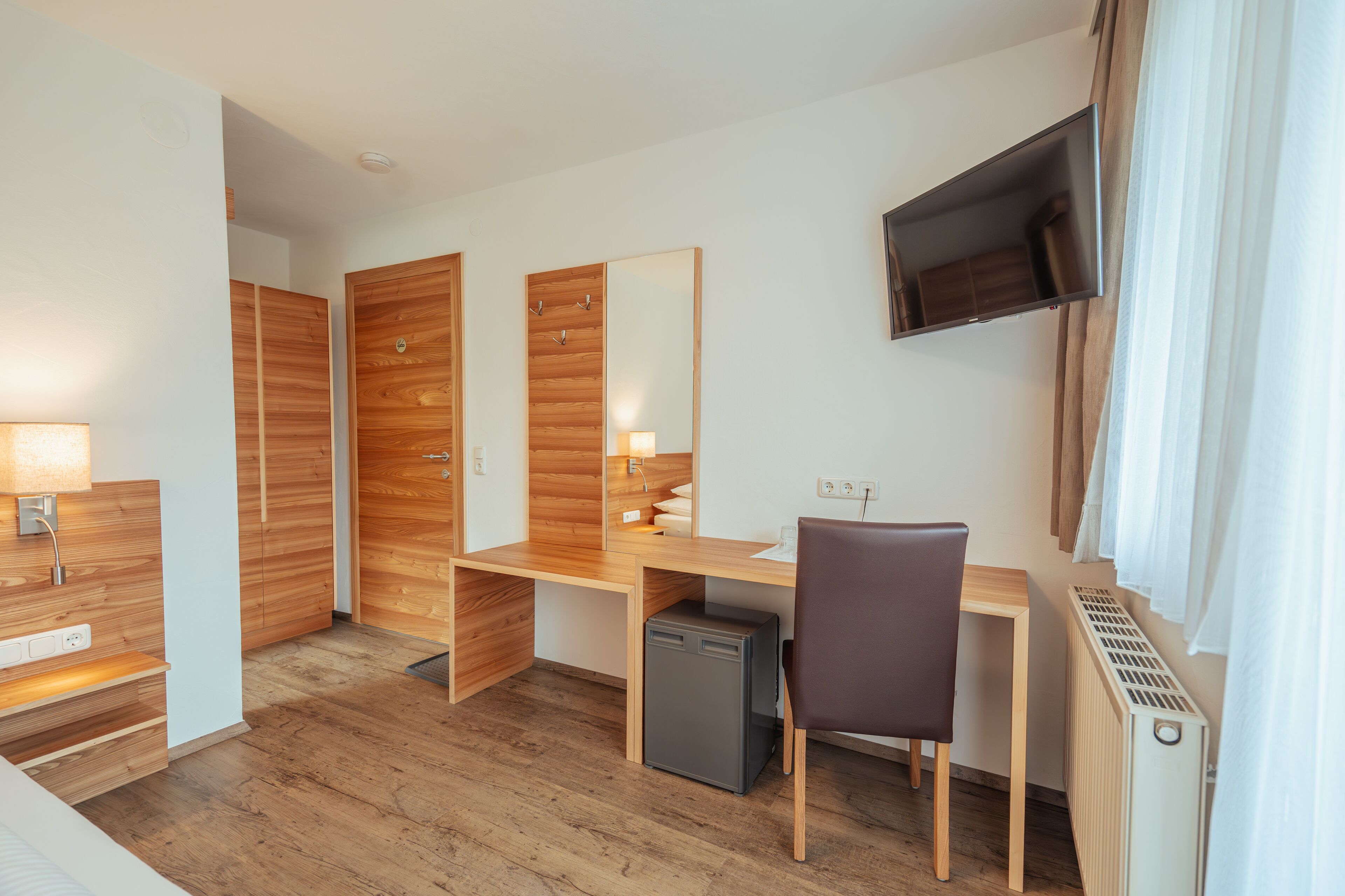 Quarto de hotel moderno com escrivaninha de madeira, uma cadeira, um frigorífico pequeno, televisão fixada na parede e janela com cortinas.
