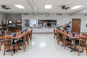 Uma sala de conferências com mesas e cadeiras de madeira, uma área de cozinha ao fundo e um relógio na parede.