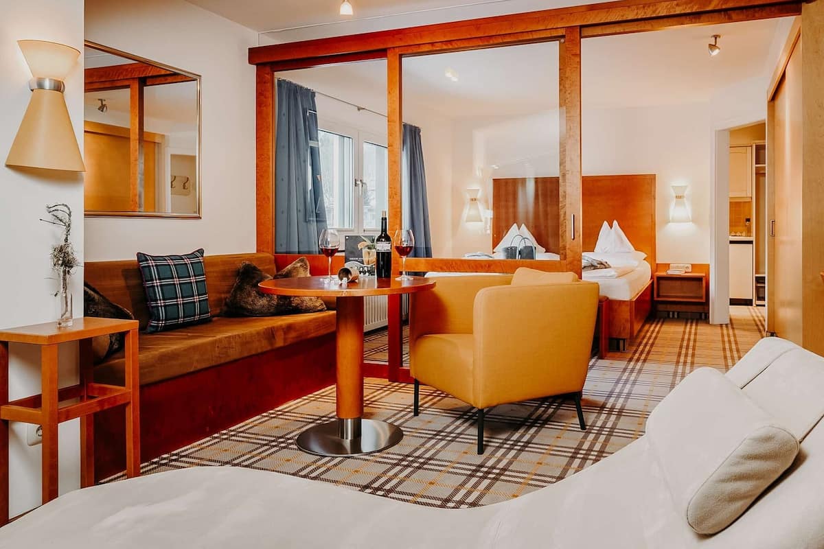 Quarto de hotel com sofá, uma pequena mesa com garrafa e copos, uma cadeira, cama e um espelho grande.