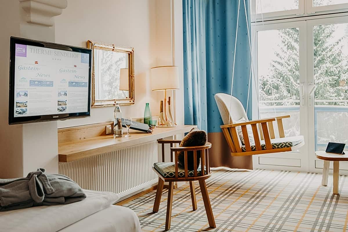 Quarto de hotel com cama, escrivaninha com computador, cadeira, janela com cortinas e vista para árvores lá fora.