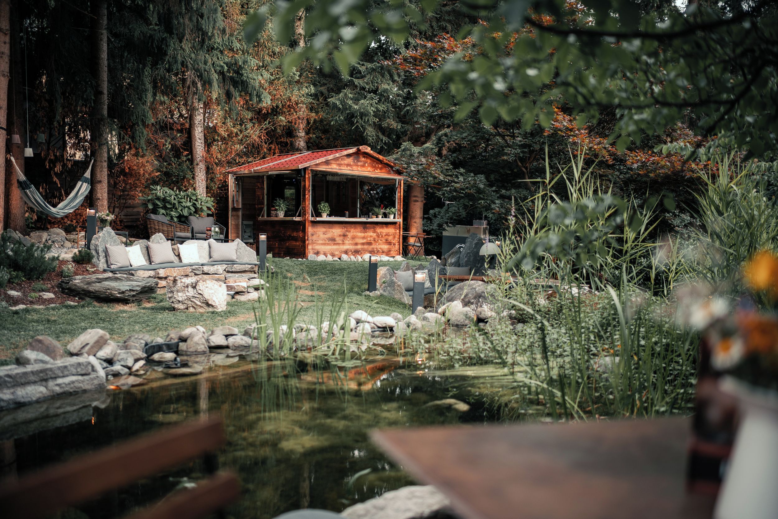 Uma cabana de madeira com estrutura de vidro, rodeada por um lago, pedras e árvores.