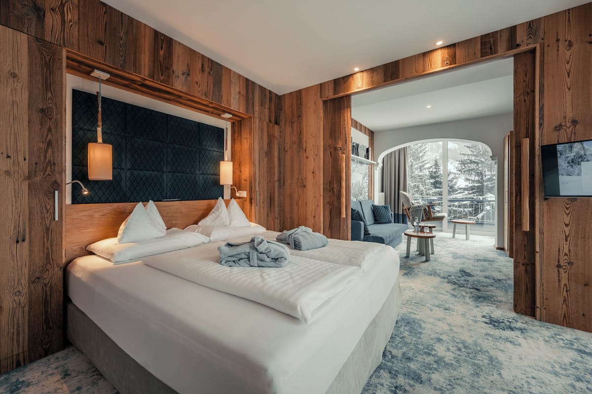Quarto de hotel moderno com uma cama grande, painéis de madeira, vista para uma paisagem nevada e uma televisão na parede.