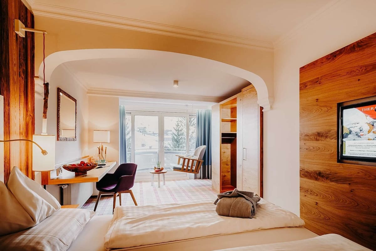 Quarto de hotel com cama, escrivaninha, cadeira, espelho e vista através de uma janela grande.