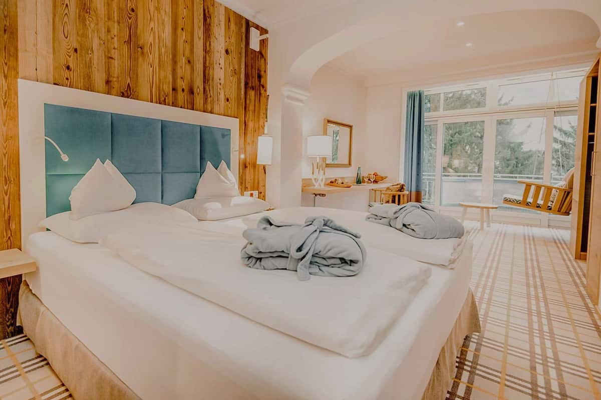 Quarto de hotel com uma cama grande, painéis de madeira nas paredes, uma mesa com luminária e vista para o exterior através de uma grande janela.