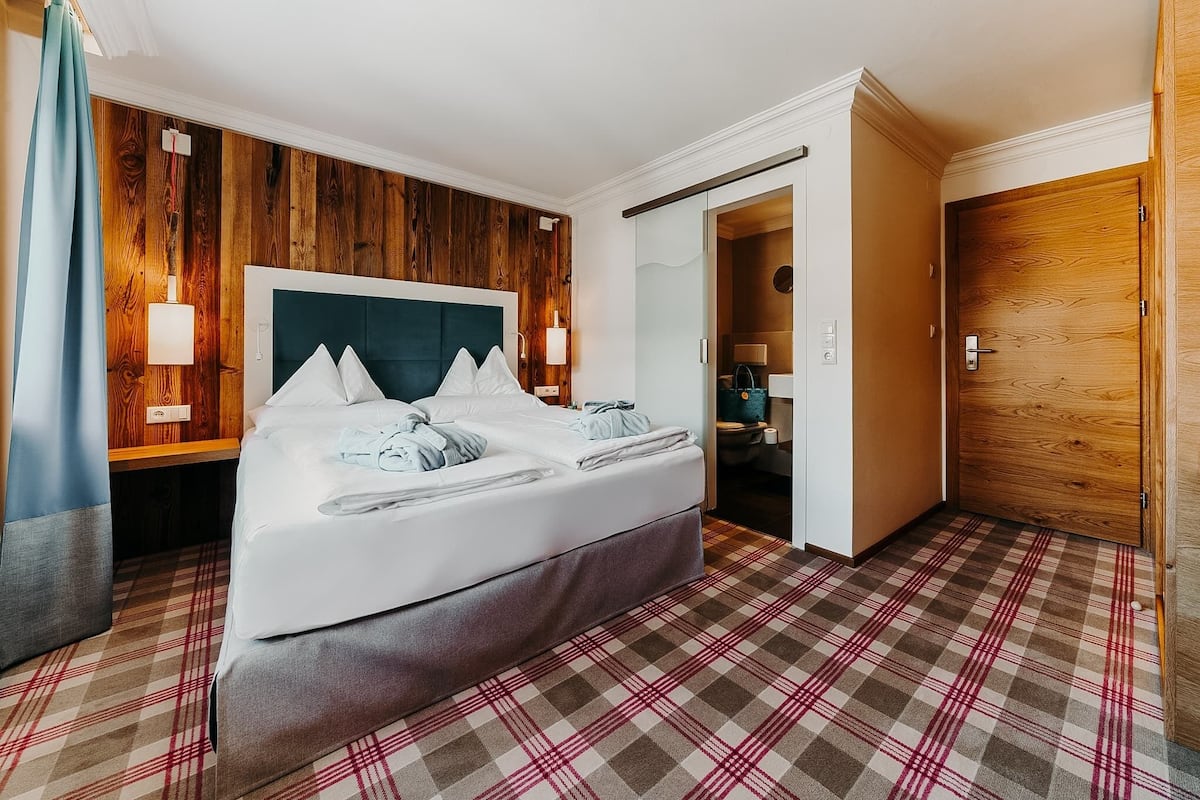 Quarto de hotel com cama, paredes de painel de madeira, chão quadriculado e porta que dá para um banheiro.