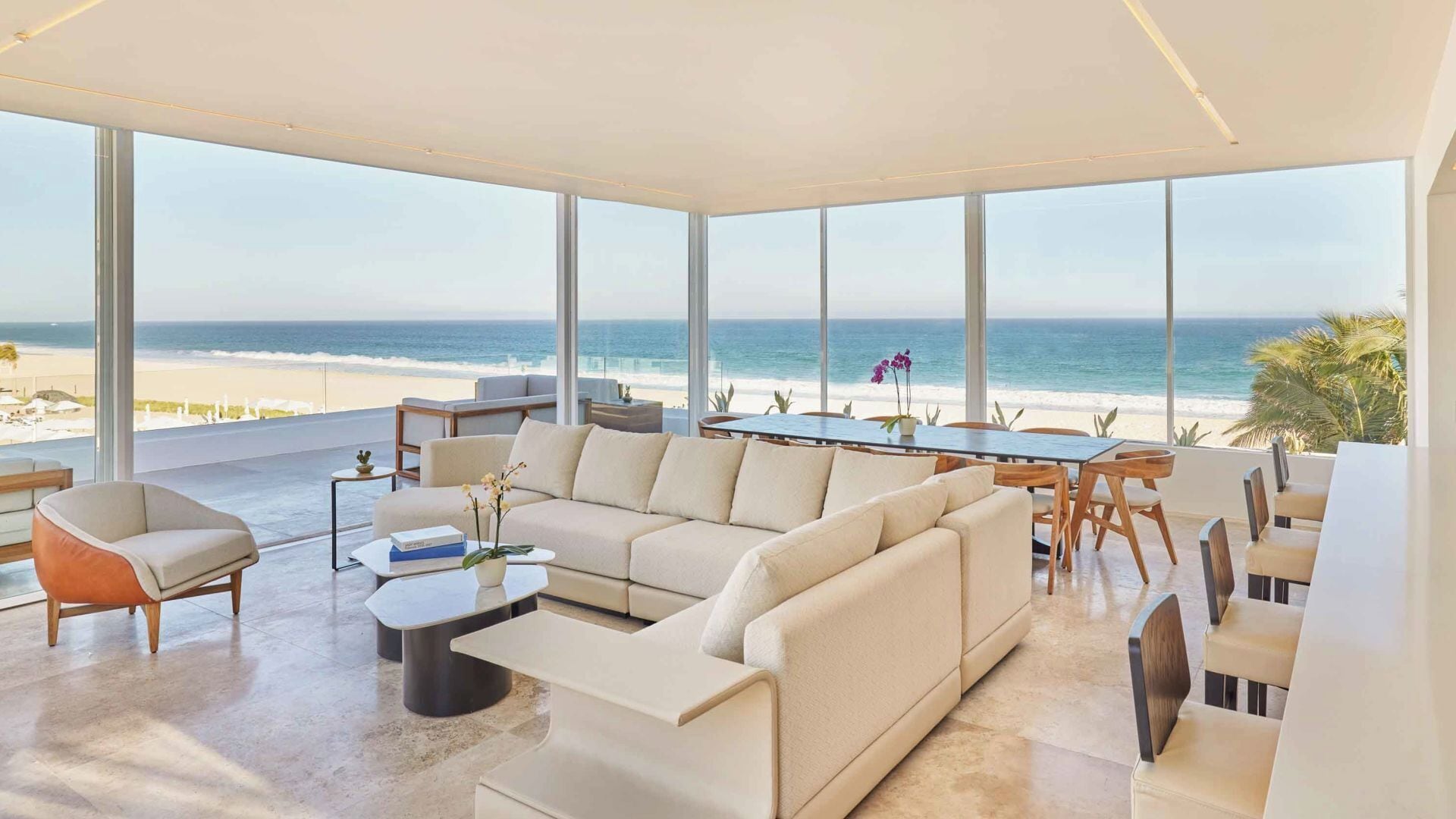 Ampla sala de estar com uma grande janela com vista para a praia. Há um sofá bege, uma mesa de centro e uma mesa de jantar com cadeiras.