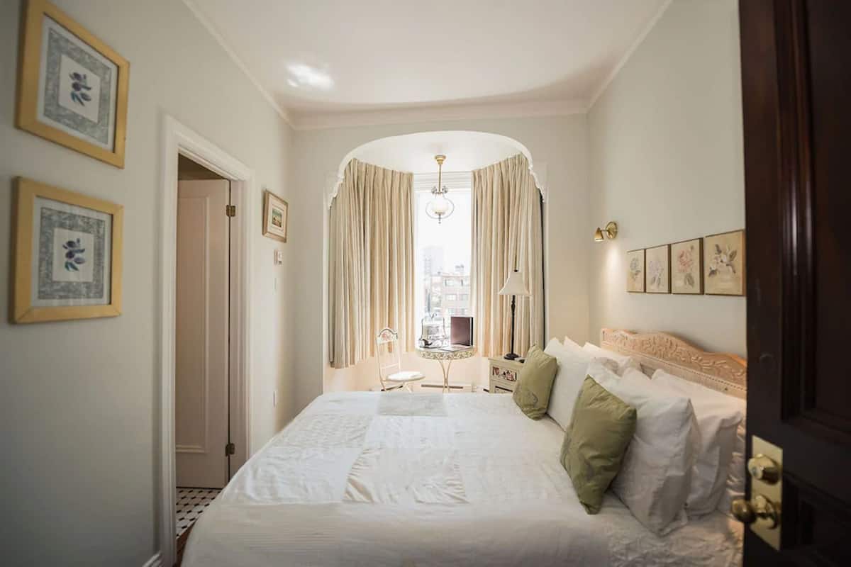 Quarto de hotel com uma cama grande, uma mesa com cadeira, vista pela janela com cortinas e quadros nas paredes.