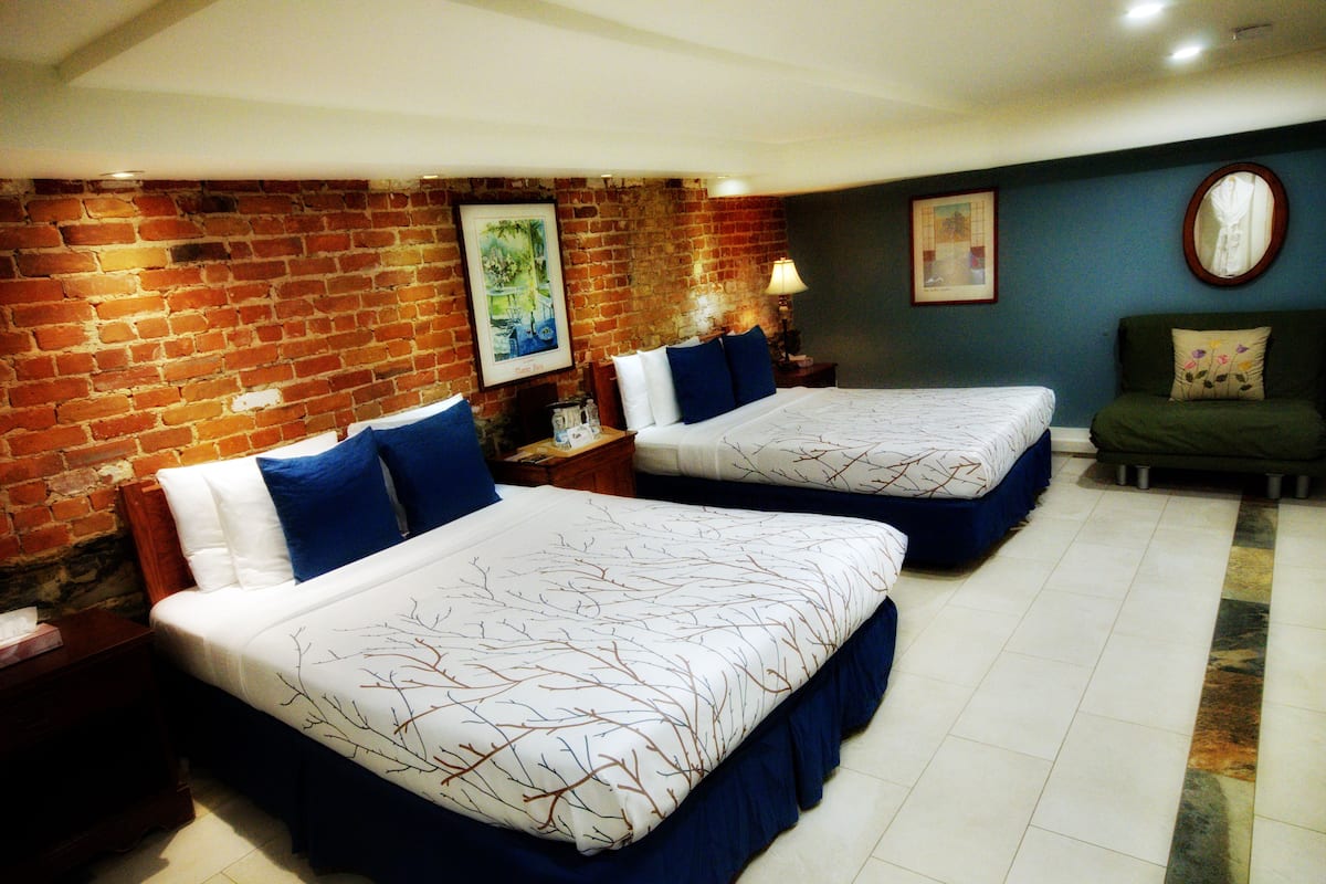 Quarto de hotel com duas camas, uma parede de tijolos, um mural, uma mesa de cabeceira e um sofá verde.