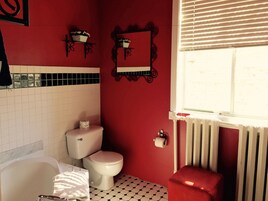 Casa de banho com parede vermelha, azulejos brancos, sanita branca, banheira branca e janela com persianas.